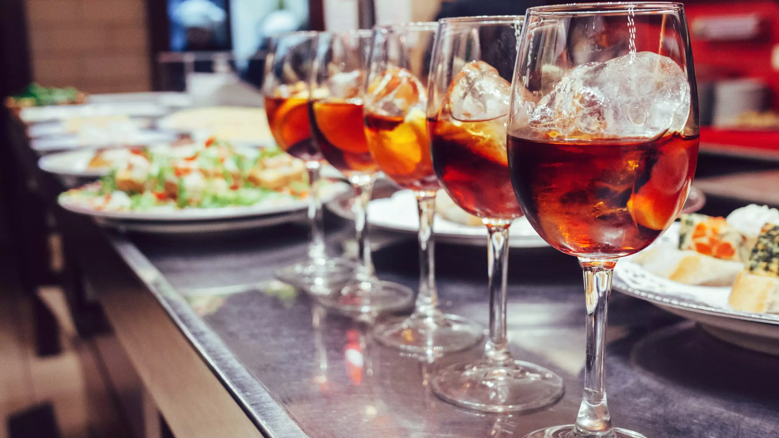 Пять стаканов вермута с кубиками льда на барной стойке рядом с несколькими тарелками тапас.